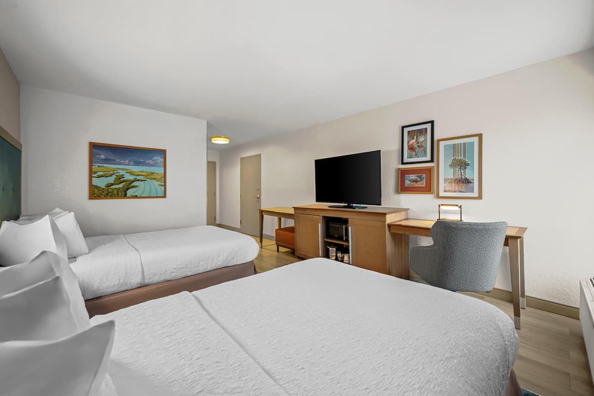 Quarto de hotel com duas camas, uma escrivaninha com televisão e obras de arte nas paredes.