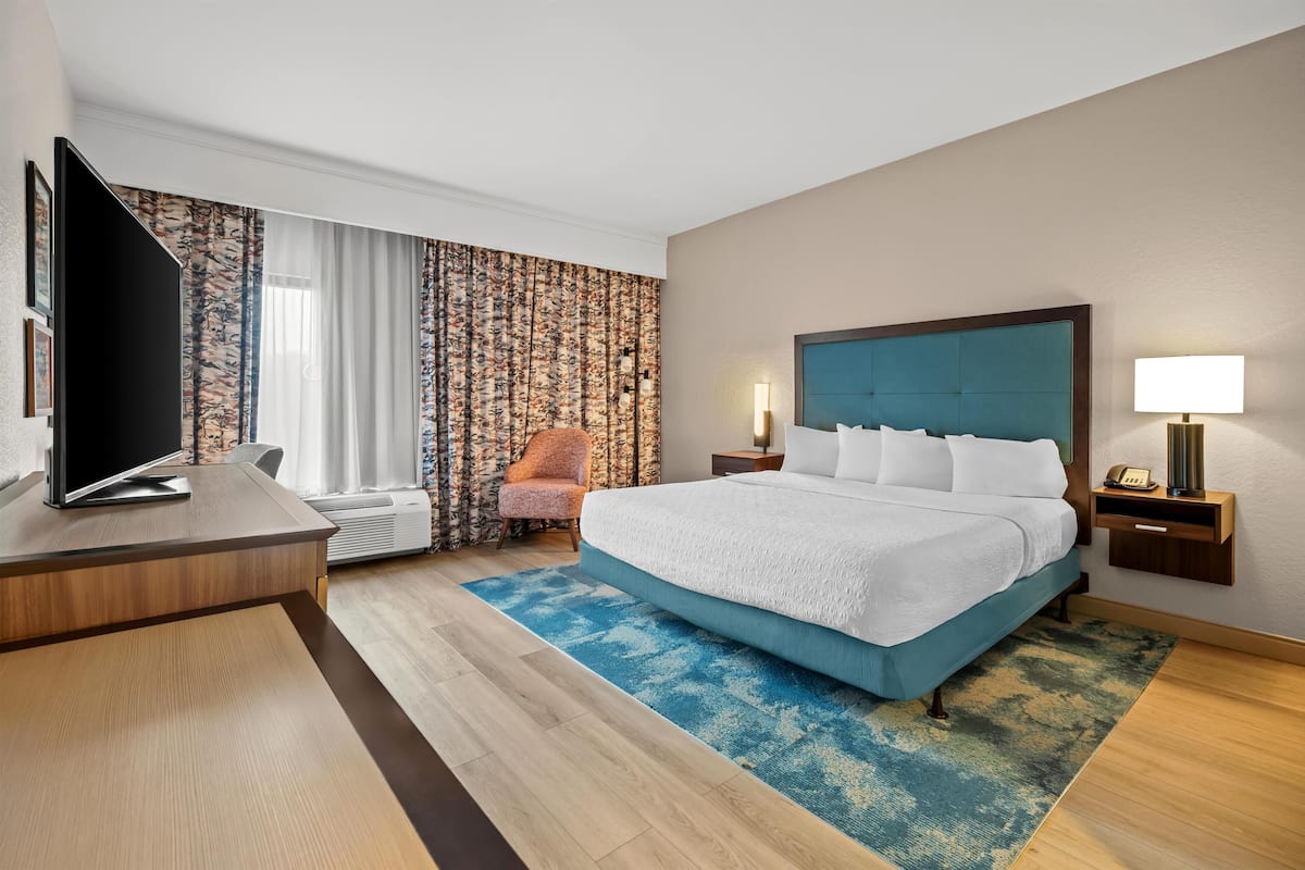 Quarto de hotel com uma cama, uma escrivaninha com televisão, uma cadeira, uma mesinha de cabeceira com abajur e uma janela com cortinas.