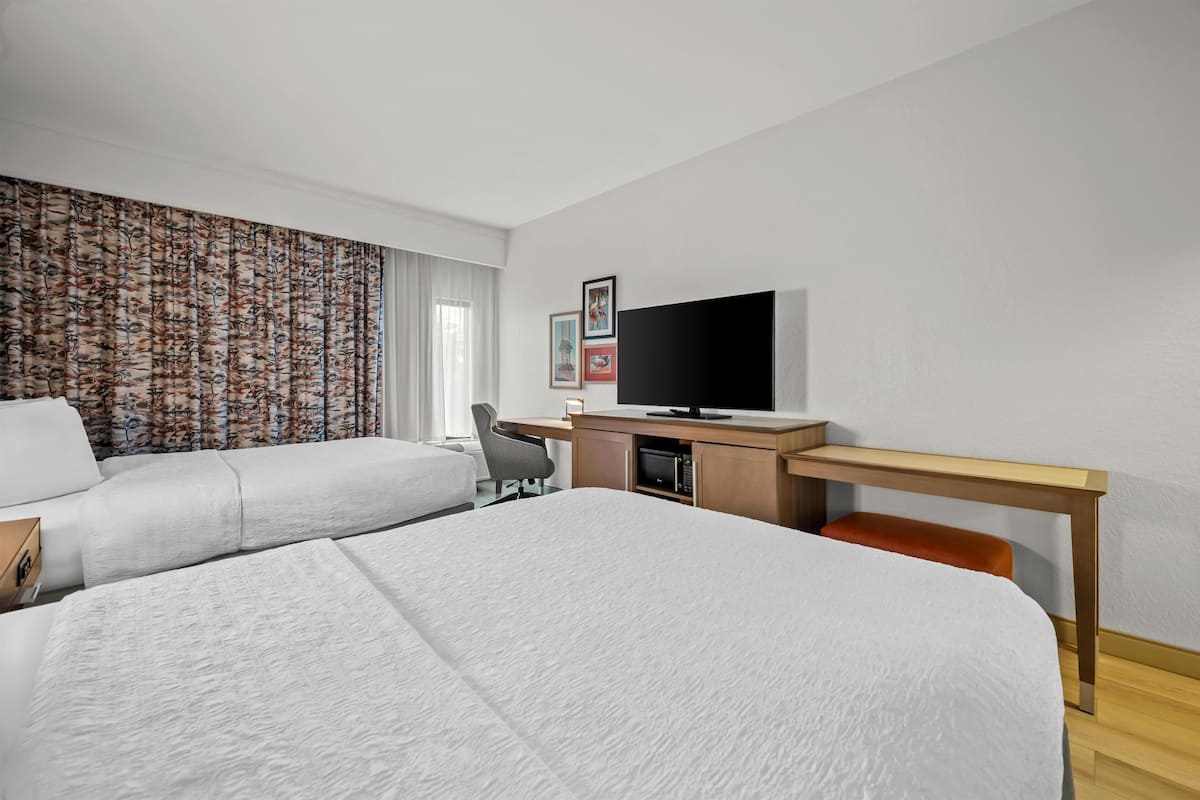 Quarto de hotel com duas camas, uma escrivaninha, televisão e janela com cortinas.