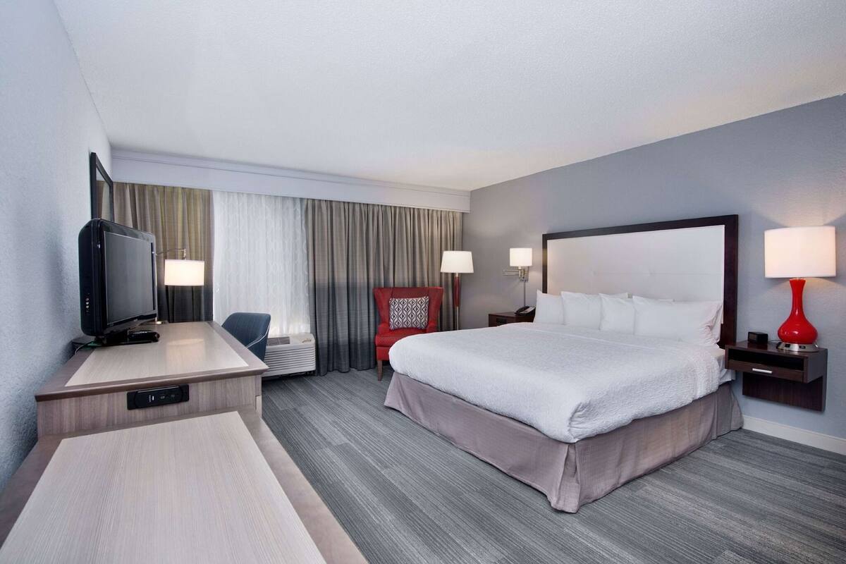 Quarto de hotel com cama, mesas de cabeceira, escrivaninha com computador, cadeira, televisão e abajur vermelho.
