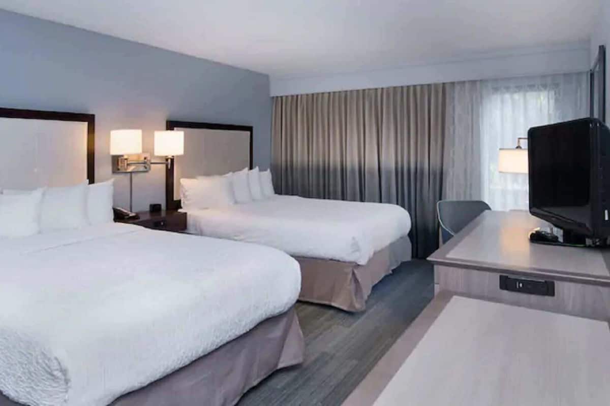 Quarto de hotel com duas camas, uma escrivaninha, televisão e uma luminária.