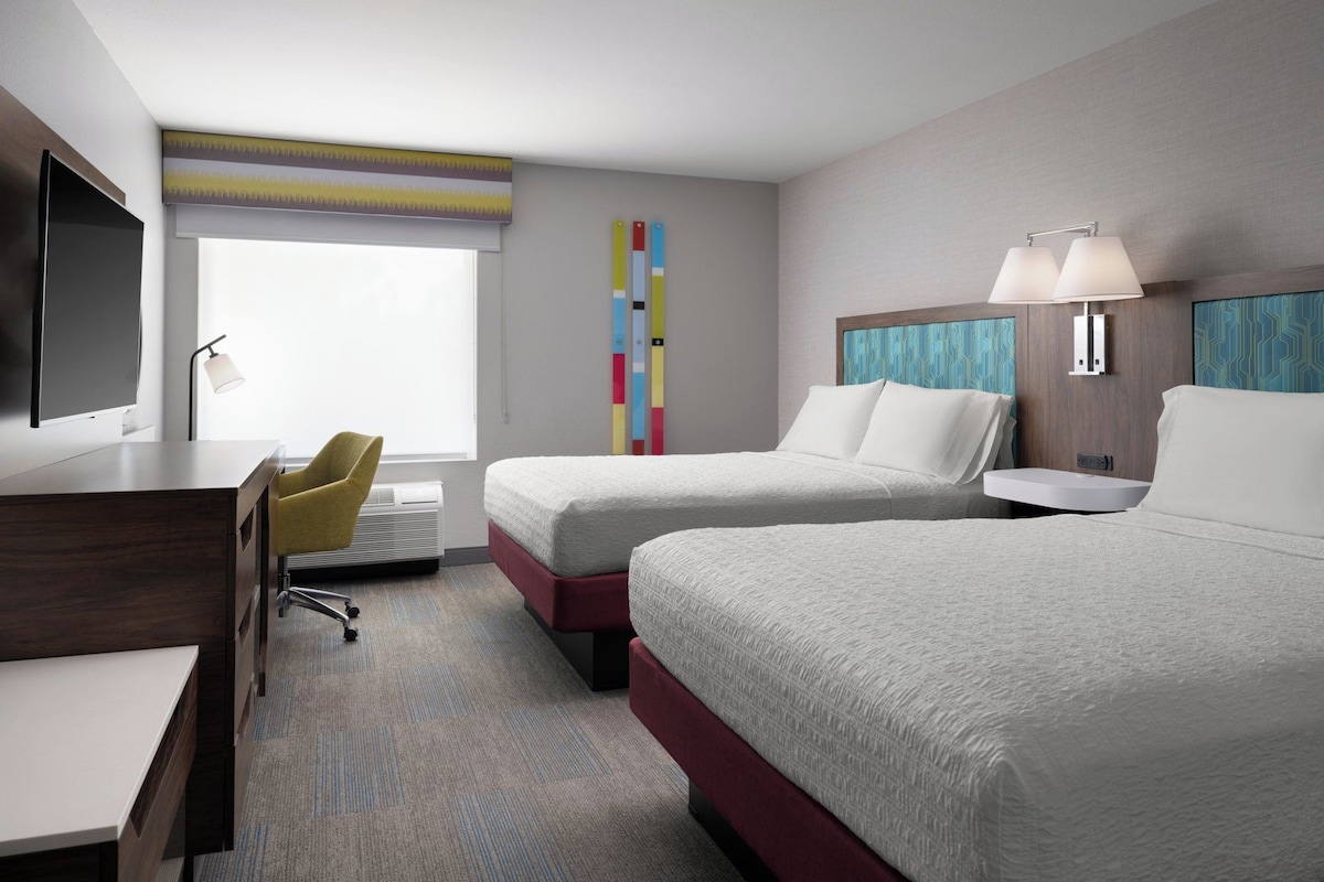 Quarto de hotel com duas camas, uma escrivaninha, uma cadeira, televisão e janela com persianas.