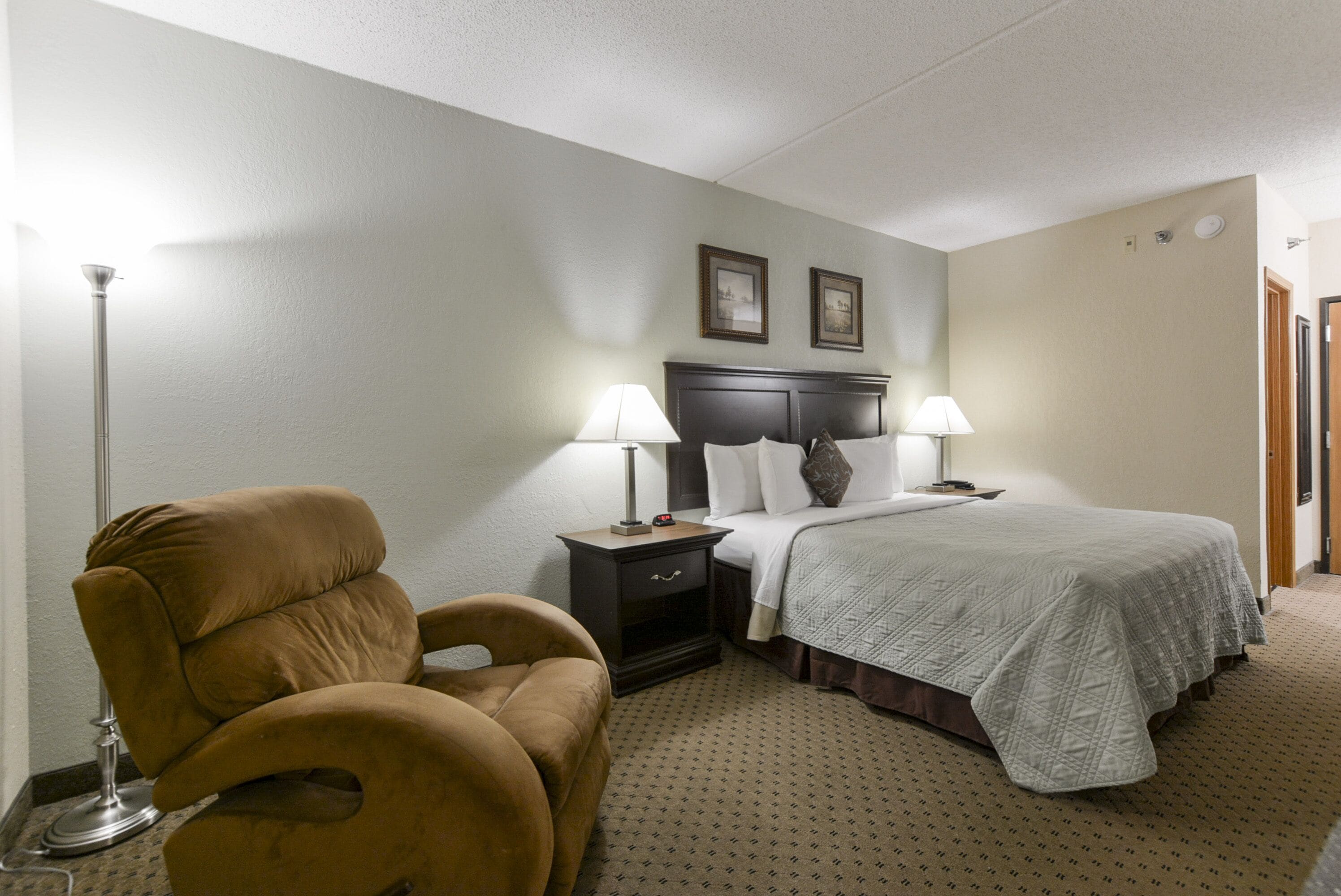 Quarto de hotel com cama, mesa de cabeceira, abajur e uma poltrona marrom.