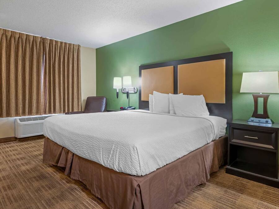 Quarto de hotel com uma cama grande, uma escrivaninha, uma cadeira, um abajur e um aquecedor fixado na parede.