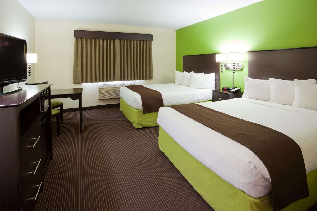 Quarto de hotel com duas camas, uma escrivaninha com televisão, uma luminária e uma janela com cortinas.
