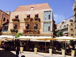 Um edifício com telhado de azulejos e várias varandas com plantas. Há mesas e cadeiras ao ar livre sob guarda-sóis, sugerindo um ambiente de café ou restaurante.