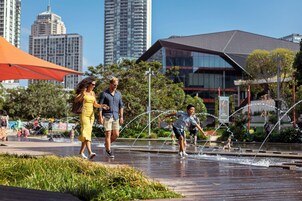 Um parque público com uma fonte, vegetação e edifícios modernos ao fundo.