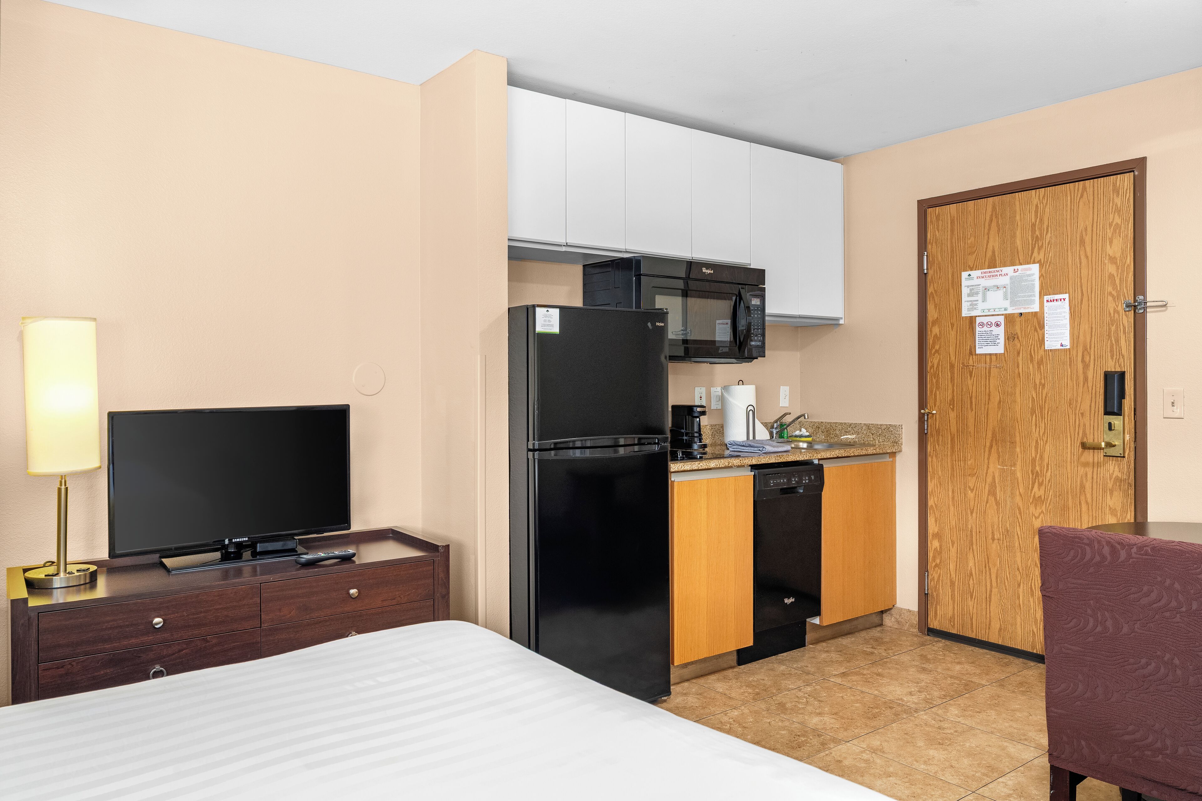 Quarto de hotel compacto com cama, televisão, frigorífico, micro-ondas, máquina de café e porta de madeira com avisos de segurança.