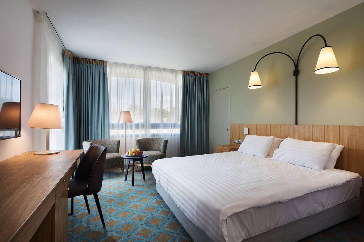 Quarto de hotel com cama, escrivaninha, cadeira, luminária e uma pequena mesa com frutas.