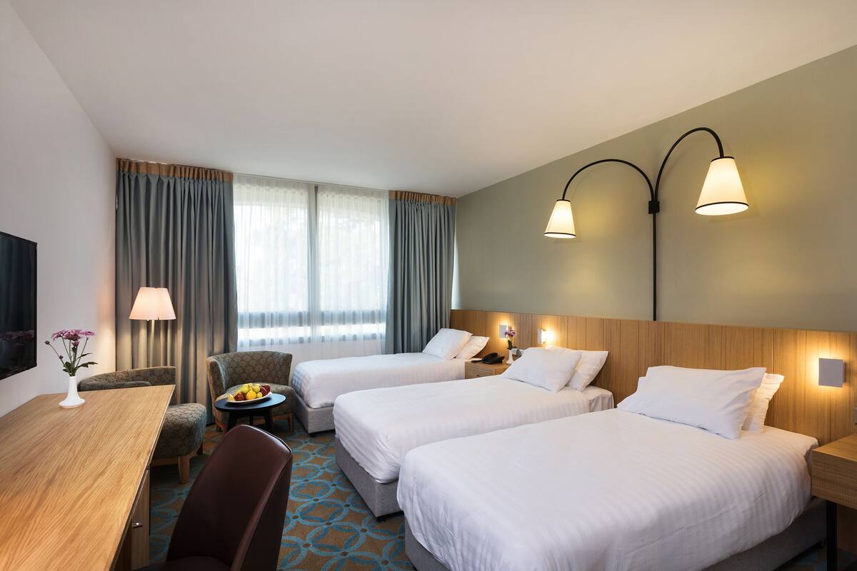 Quarto de hotel com duas camas, uma mesa, uma cadeira, uma mesinha com fruta e um abajur.