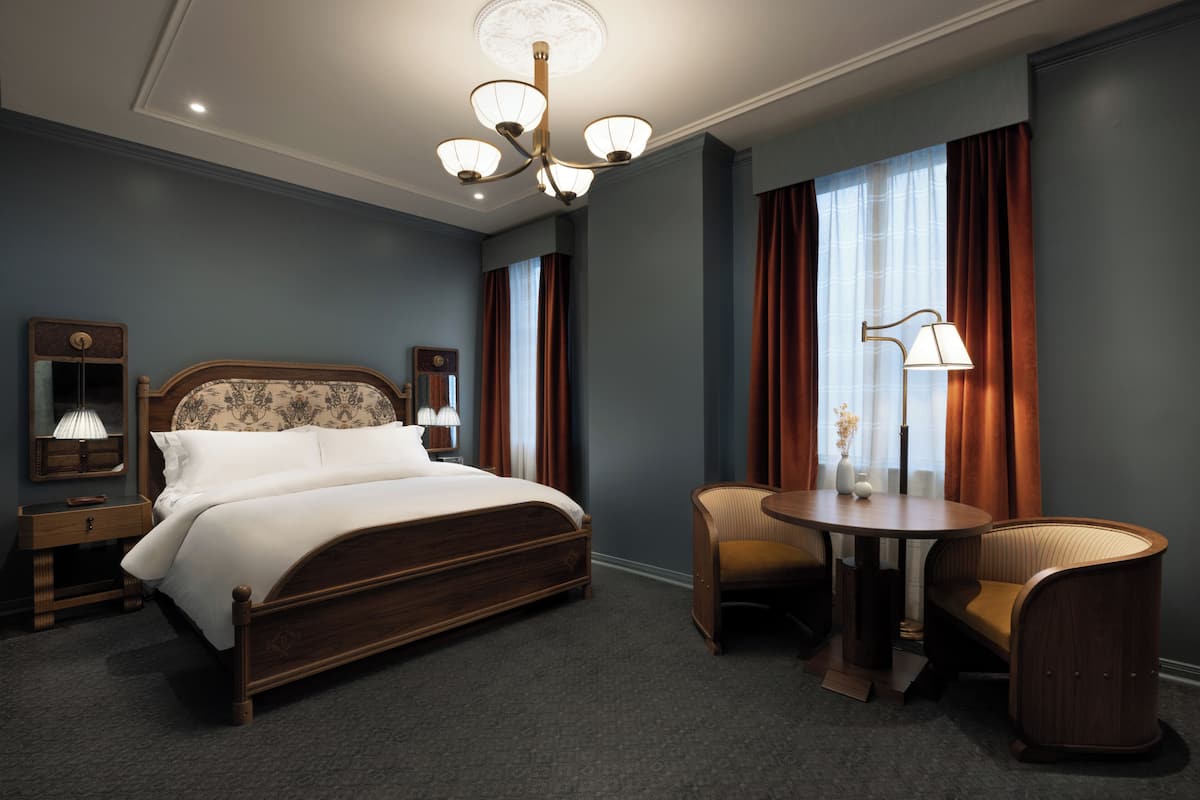 Quarto de hotel com uma cama grande, uma mesa redonda, duas cadeiras, uma mesinha de cabeceira, um abajur e um espelho.