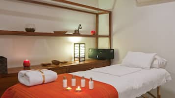 Uma cama bem arrumada com lençóis brancos e um cobertor listrado vermelho, uma pequena mesa com uma tigela e velas, e uma estante com um espelho e objetos decorativos.