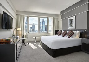 Quarto de hotel moderno com uma cama grande, uma escrivaninha com TV de tela plana, uma cadeira e vista para o horizonte urbano através de grandes janelas.