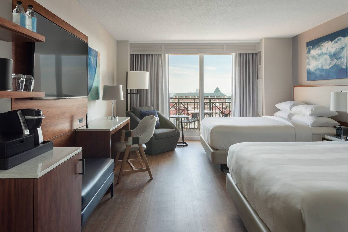 Quarto de hotel com secretária, duas camas, uma cadeira, televisão e varanda com vista.
