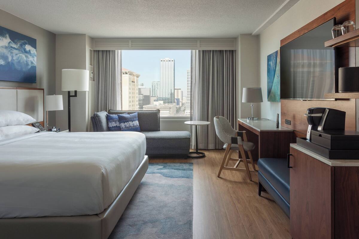 Quarto de hotel com uma cama grande, uma escrivaninha, uma cadeira, uma mesinha redonda e vista para a paisagem urbana pela janela.