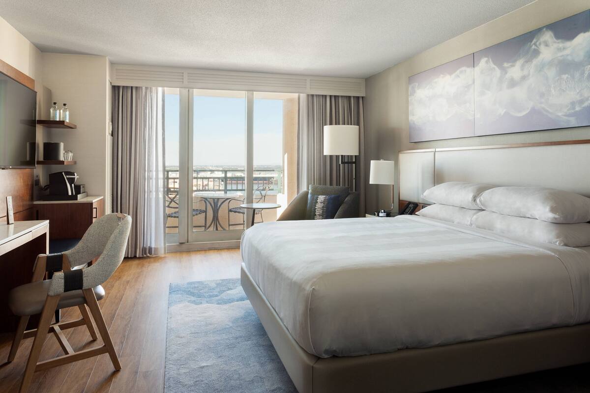 Quarto de hotel com uma cama grande, uma escrivaninha com cadeira, televisão e vista para a cidade pela varanda.