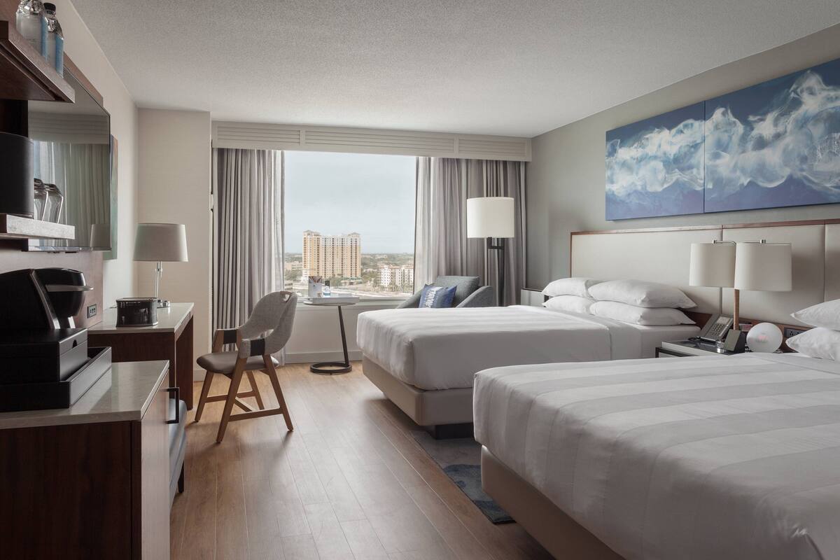 Quarto de hotel com duas camas, uma escrivaninha com computador, uma cadeira e vista da cidade por grandes janelas.