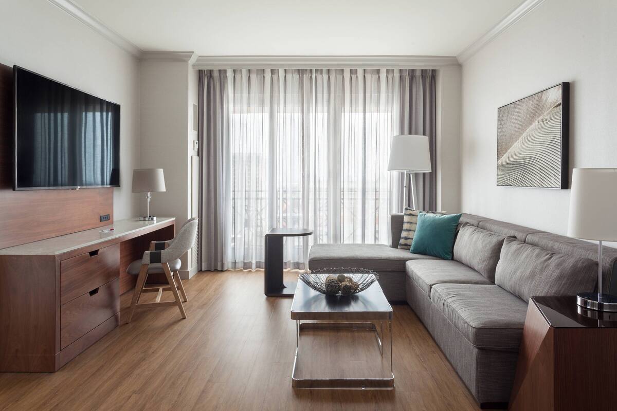 Uma sala de estar moderna com televisão de tela plana, uma escrivaninha com gavetas, um sofá com almofada, uma mesa de centro e uma janela grande com cortinas leves.