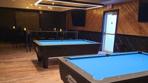Uma mesa de bilhar com superfície de feltro azul e um logotipo, situada em um espaço com piso de madeira e teto inclinado.