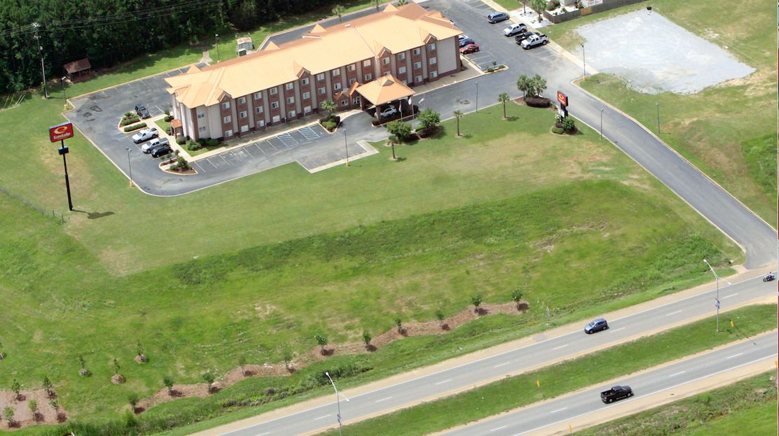 Vista aérea de um hotel com estacionamento, uma estrada e área gramada com árvores pequenas.