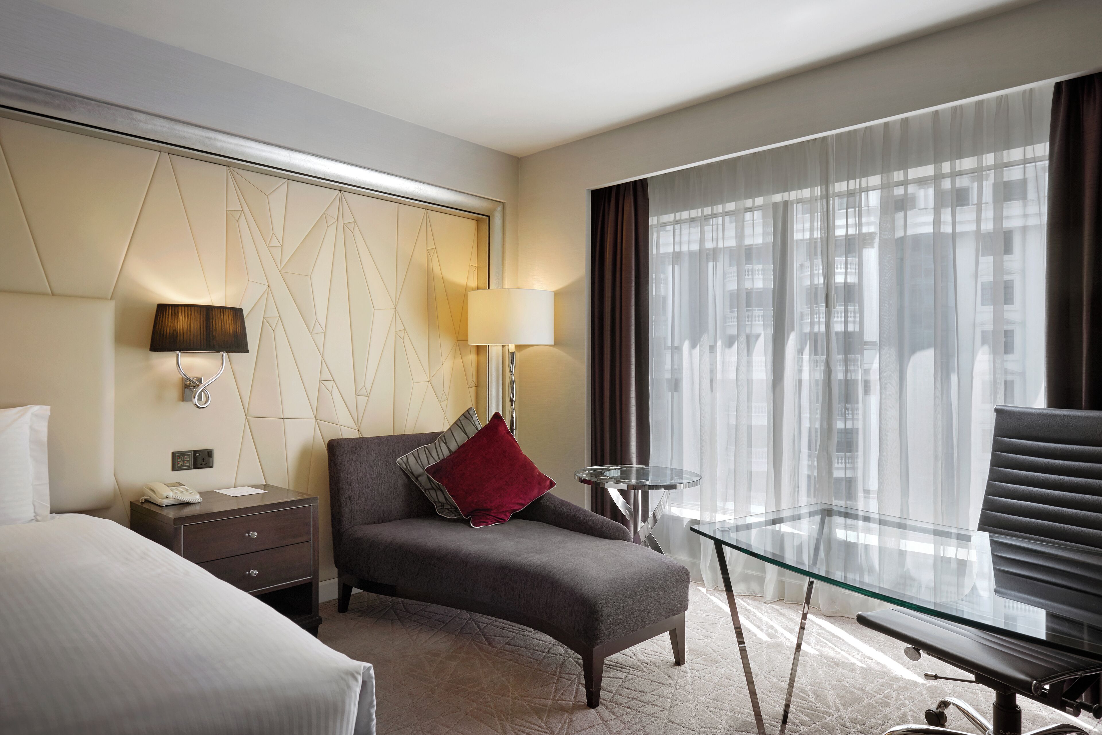 Quarto de hotel com cama, sofá, mesa de vidro, luminária e vista da cidade por grandes janelas.
