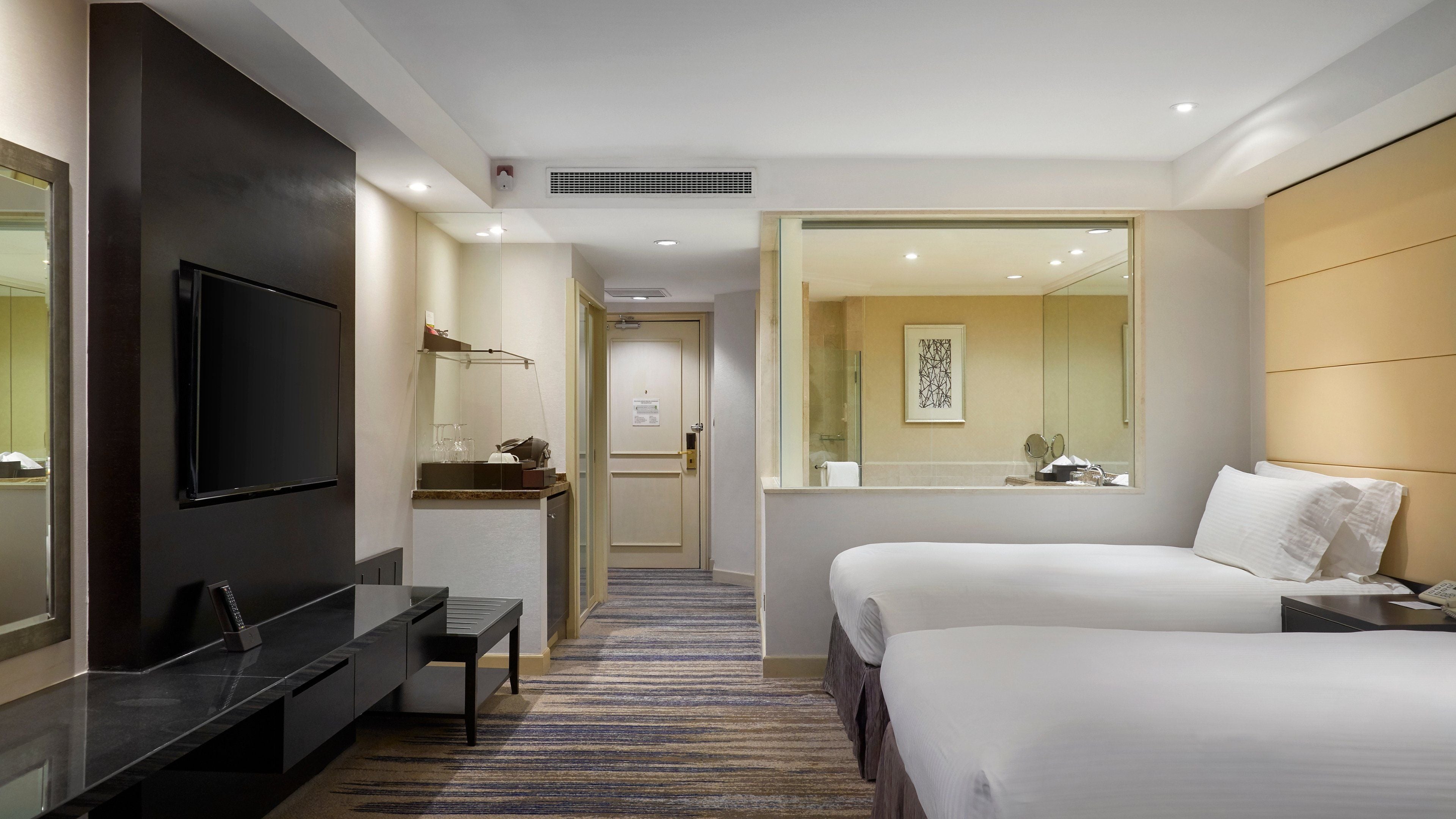 Quarto de hotel com televisão, secretária com cadeira, cama, banheiro com espelho e piso coberto por carpete.