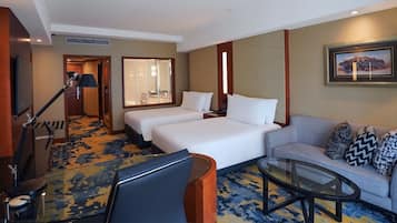 Quarto de hotel com duas camas, um sofá, mesa de centro de vidro, televisão e banheiro com chuveiro e vaso sanitário.