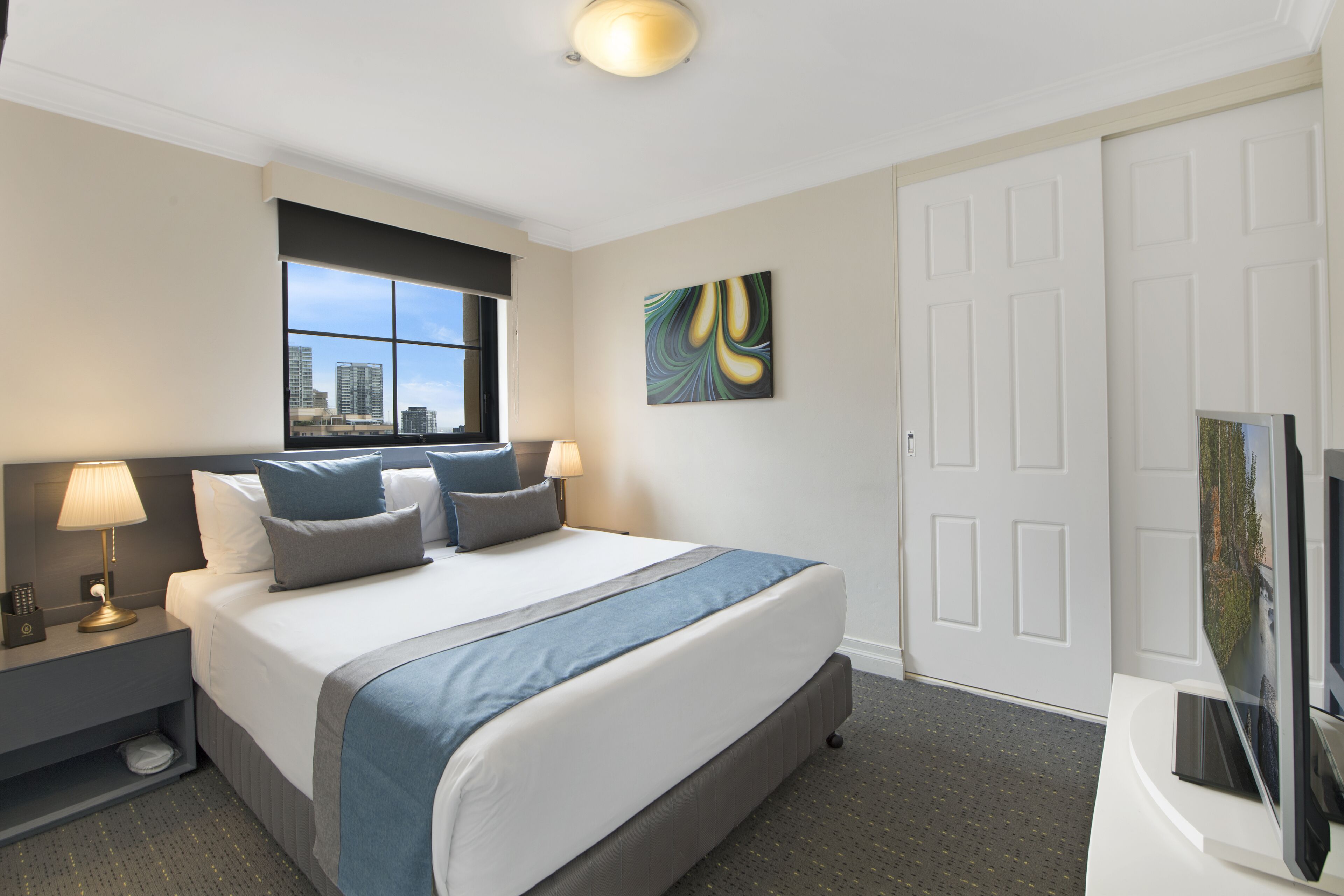 Quarto de hotel com uma cama grande, mesa de cabeceira com abajur, janela com vista para a cidade, porta e um quadro na parede.