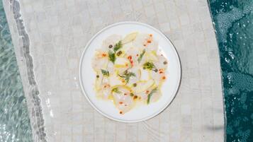 Prato branco com prato de frutos do mar, decorado com endro e alcaparras, apoiado em superfície azulejada perto de uma piscina.