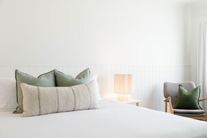 Cama bem arrumada com travesseiros, abajur e uma cadeira em quarto com paredes brancas e cabeceira de madeira.