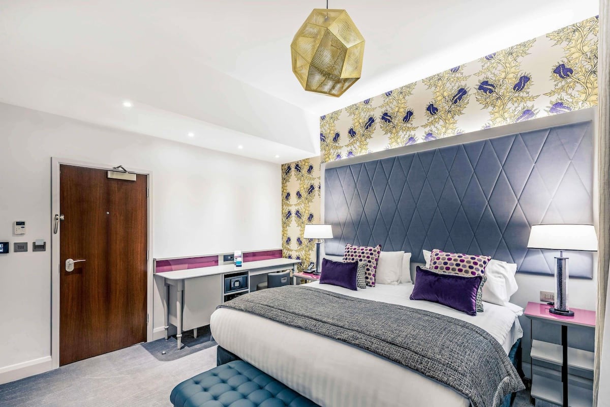Quarto de hotel com cama, escrivaninha, banqueta, papel de parede estampado e luminária geométrica.
