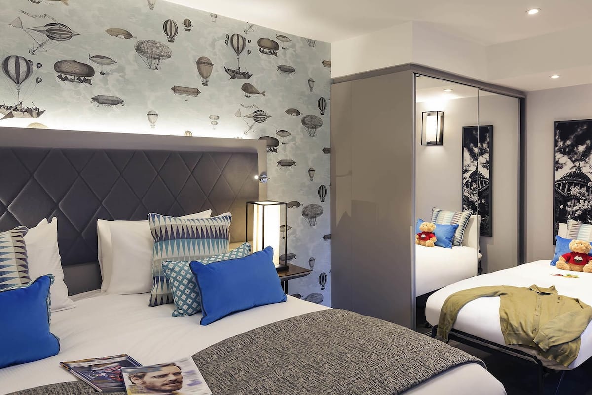 Quarto de hotel com uma cama grande, uma cama menor, uma mesinha de cabeceira, um abajur e uma parede com um padrão de balões de ar quente.