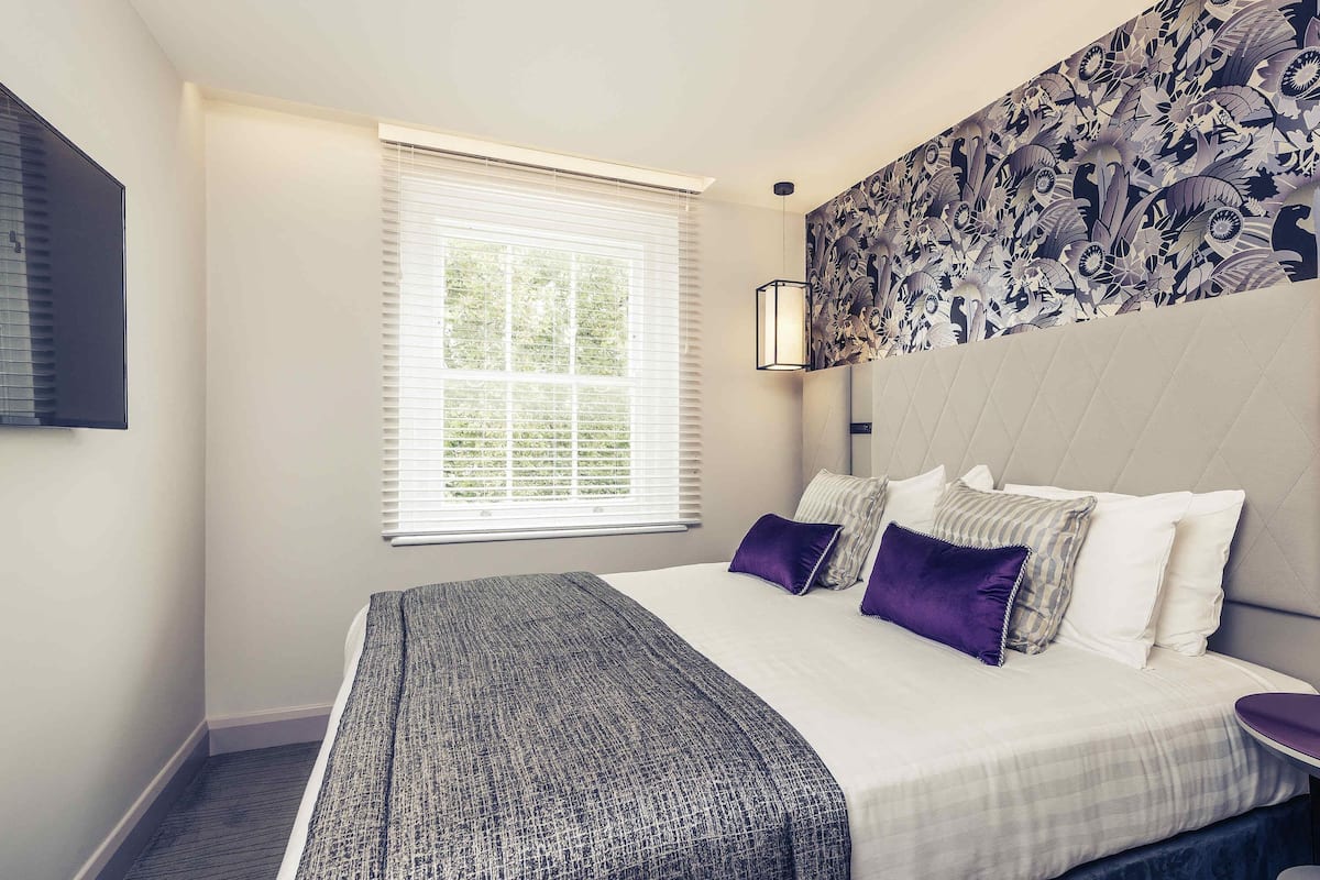 Quarto de hotel moderno com uma cama grande, cabeceira estampada, janela com persianas e uma obra de arte na parede.