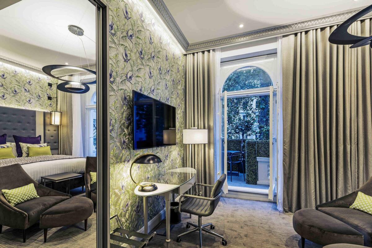 Quarto de hotel com cama, escrivaninha, duas poltronas, televisão e uma janela grande com cortinas.