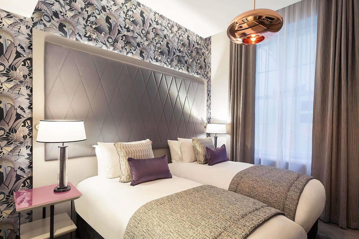 Quarto de hotel com uma cama grande, duas mesas de cabeceira, um candelabro moderno e papel de parede estampado.