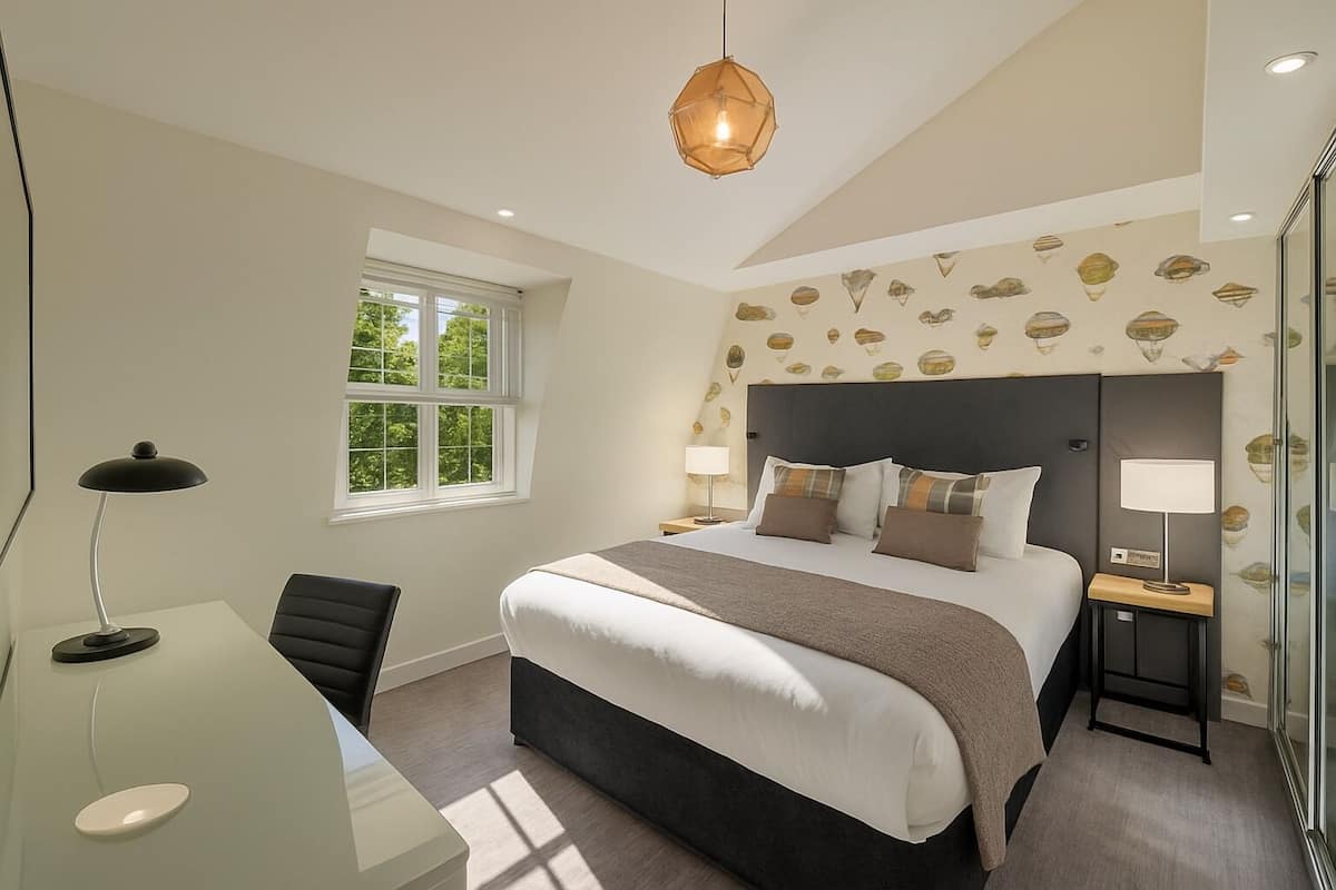 Quarto moderno com cama grande, escrivaninha e cadeira. Uma janela oferece iluminação natural e o ambiente apresenta cores neutras.