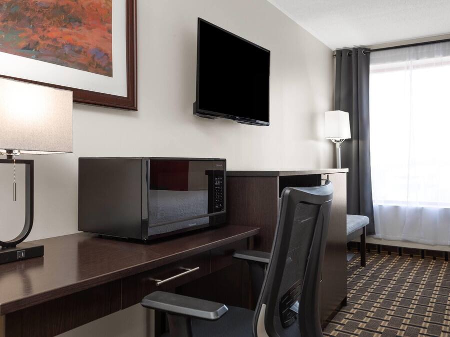 Quarto de hotel com televisão de tela plana na parede, micro-ondas numa escrivaninha de madeira, uma cadeira, luminária de mesa e janela com cortinas.