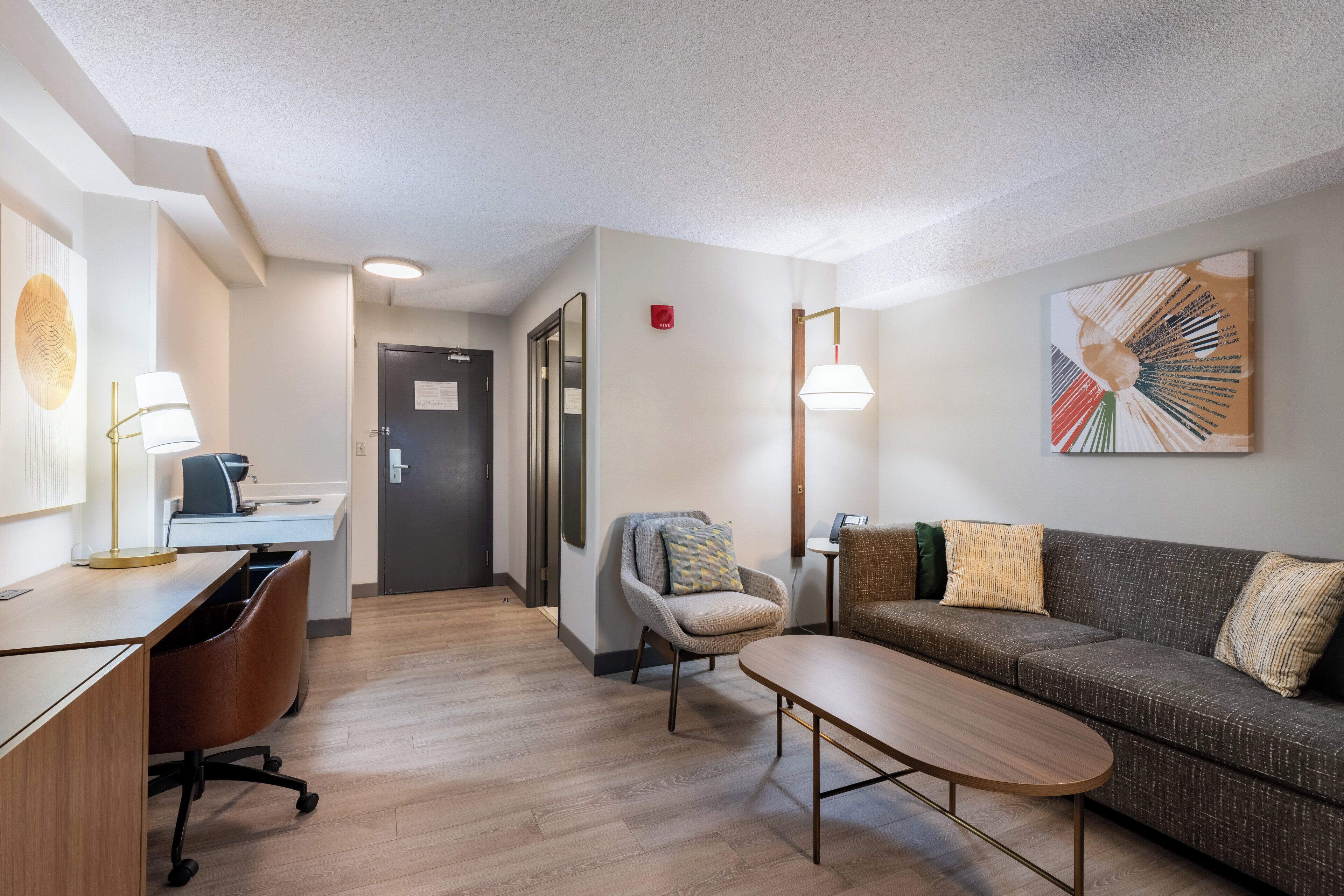 Quarto de hotel moderno com escrivaninha de madeira, sofá confortável, mesinha e luminária fixada na parede.