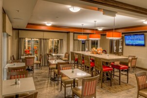 Uma área de refeições de hotel com várias mesas e cadeiras, um bar central e uma televisão na parede.