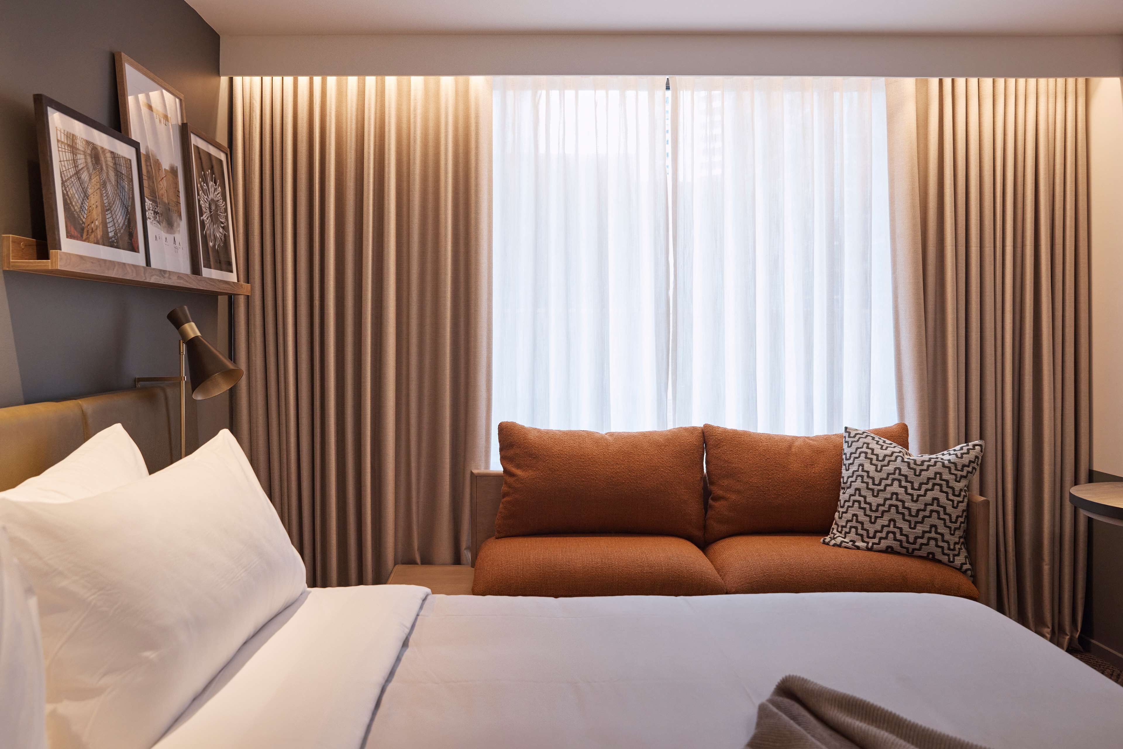 Quarto de hotel com uma cama grande, um sofá com almofadas, uma mesa de cabeceira e uma janela com cortinas.