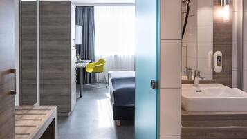Quarto de hotel com cama, escrivaninha com cadeira, banheiro com pia e espelho e janela com cortinas.