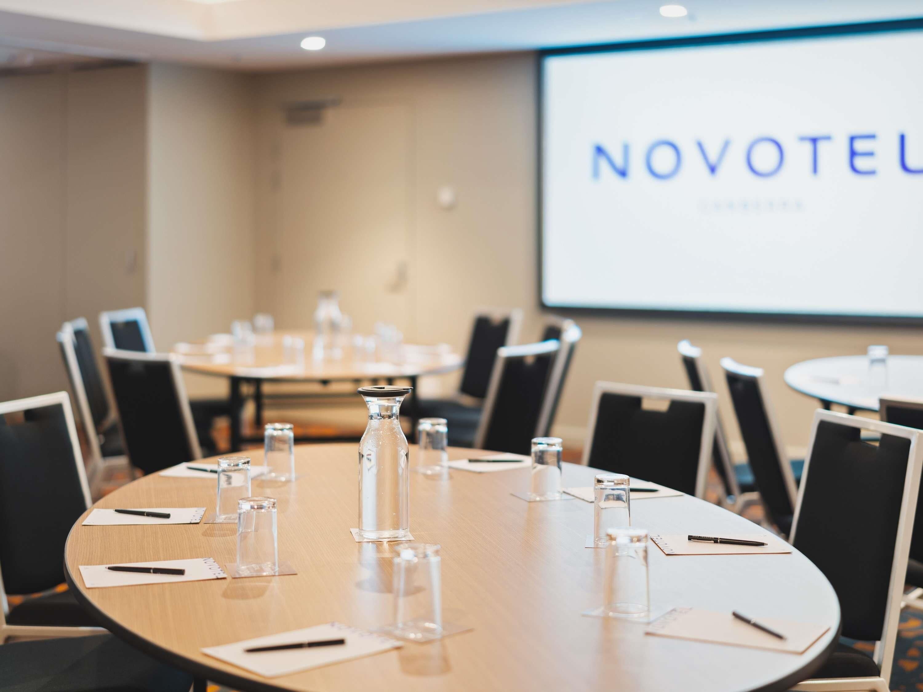 Uma sala de conferências com uma tela grande exibindo 'NOVOTEL' e uma longa mesa com cadeiras, copos de água e canetas.