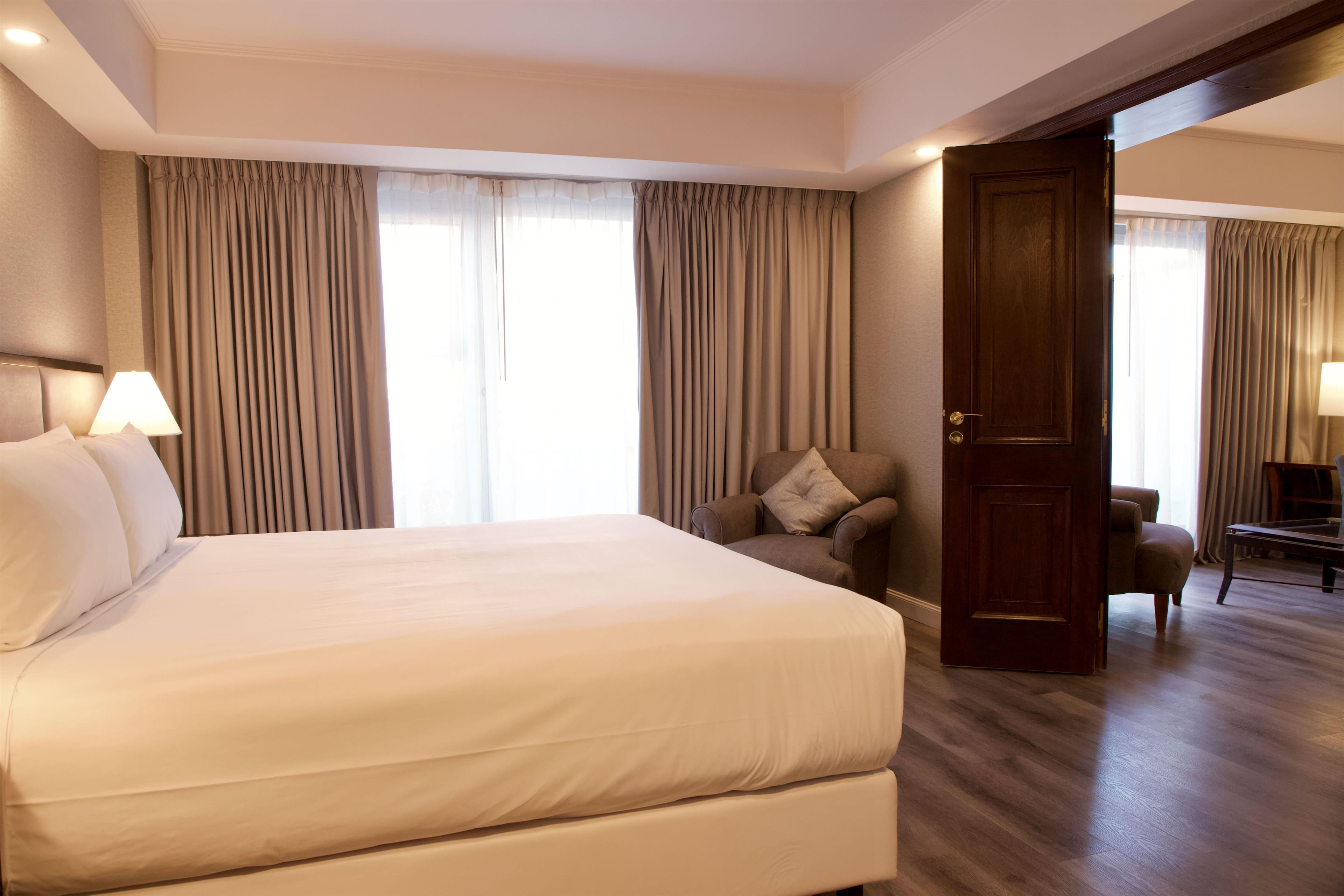 Quarto de hotel com uma cama grande, duas cadeiras, uma porta de madeira e cortinas.