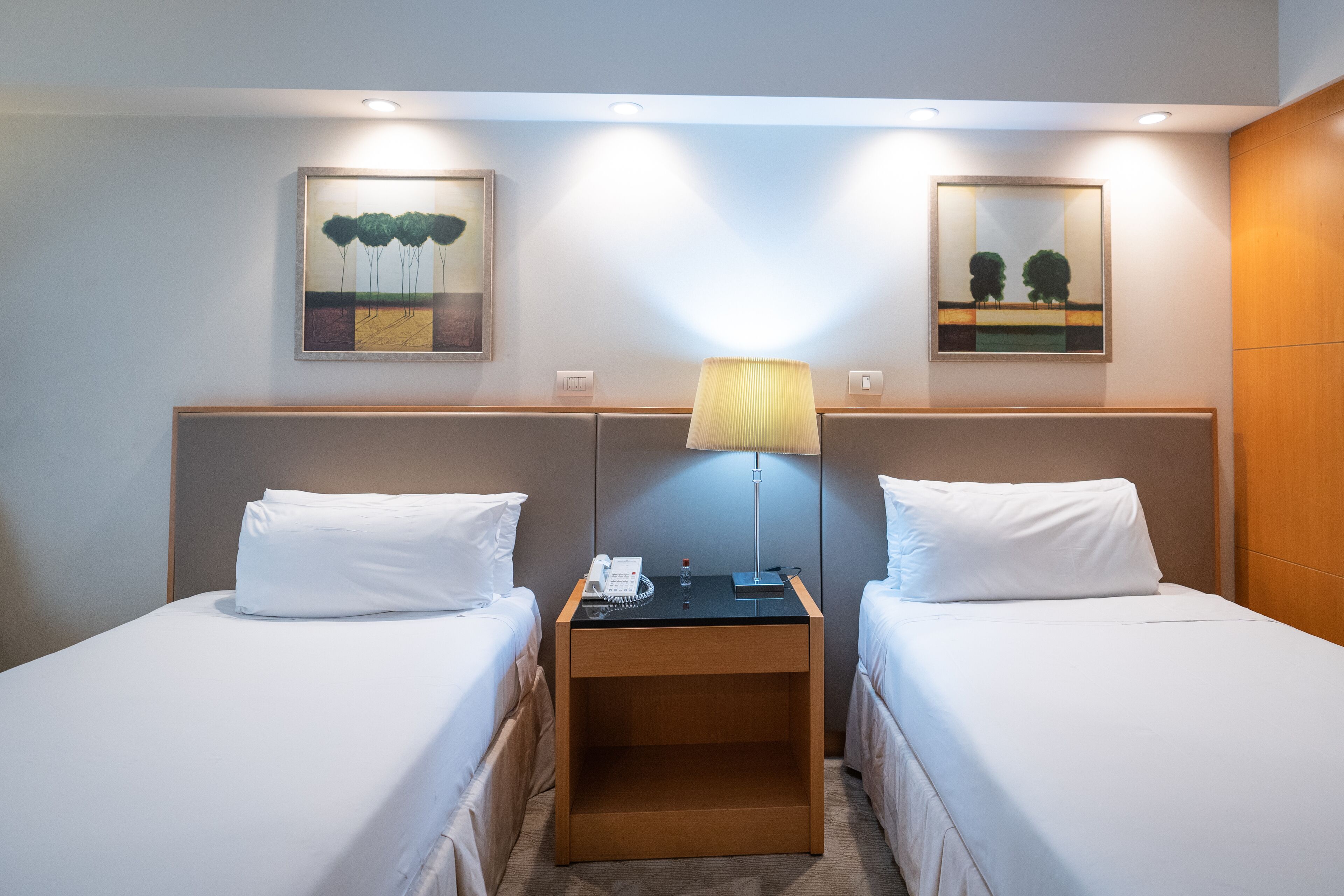 Quarto de hotel com duas camas de solteiro, mesa de cabeceira com abajur, telefone e obras de arte nas paredes.