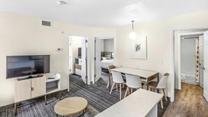 Quarto de hotel moderno com televisão de tela plana, área de refeições com mesa e cadeiras, uma pequena mesa redonda e área de chuveiro com porta de vidro.
