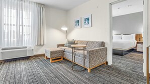 Quarto de hotel moderno com sofá cinzento, uma pequena mesa redonda, cama com roupa de cama branca, mesa de cabeceira e uma grande janela com cortinas translúcidas.