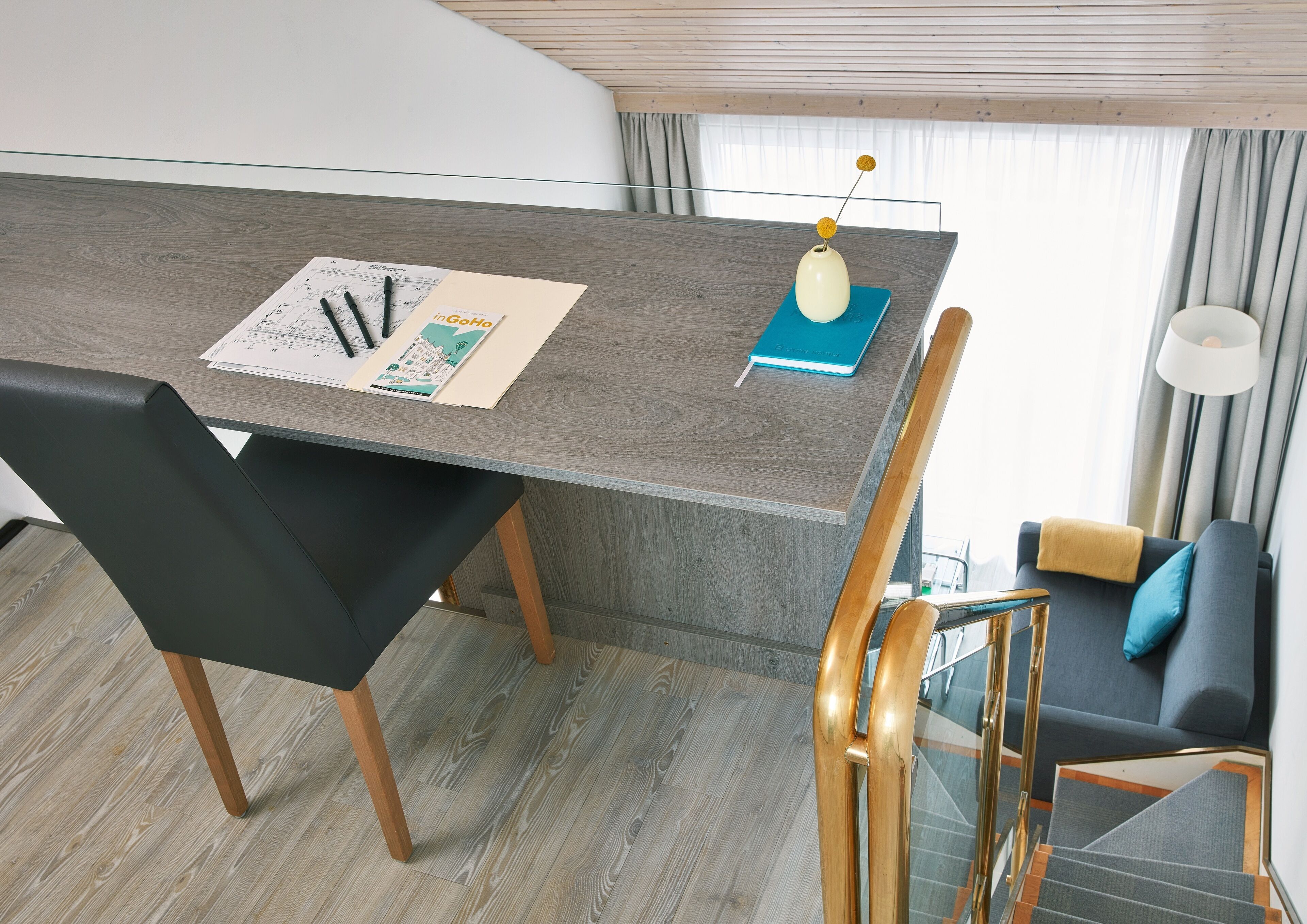 Quarto de hotel moderno com piso de madeira, uma mesa de jantar cinza com um vaso e um caderno, uma cadeira preta, um sofá com almofadas e uma escada com corrimão de vidro.