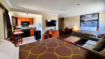 Quarto de hotel com cozinha compacta, televisão, cama com pôster de vista para montanhas e área de estar separada com mesa e cadeiras.