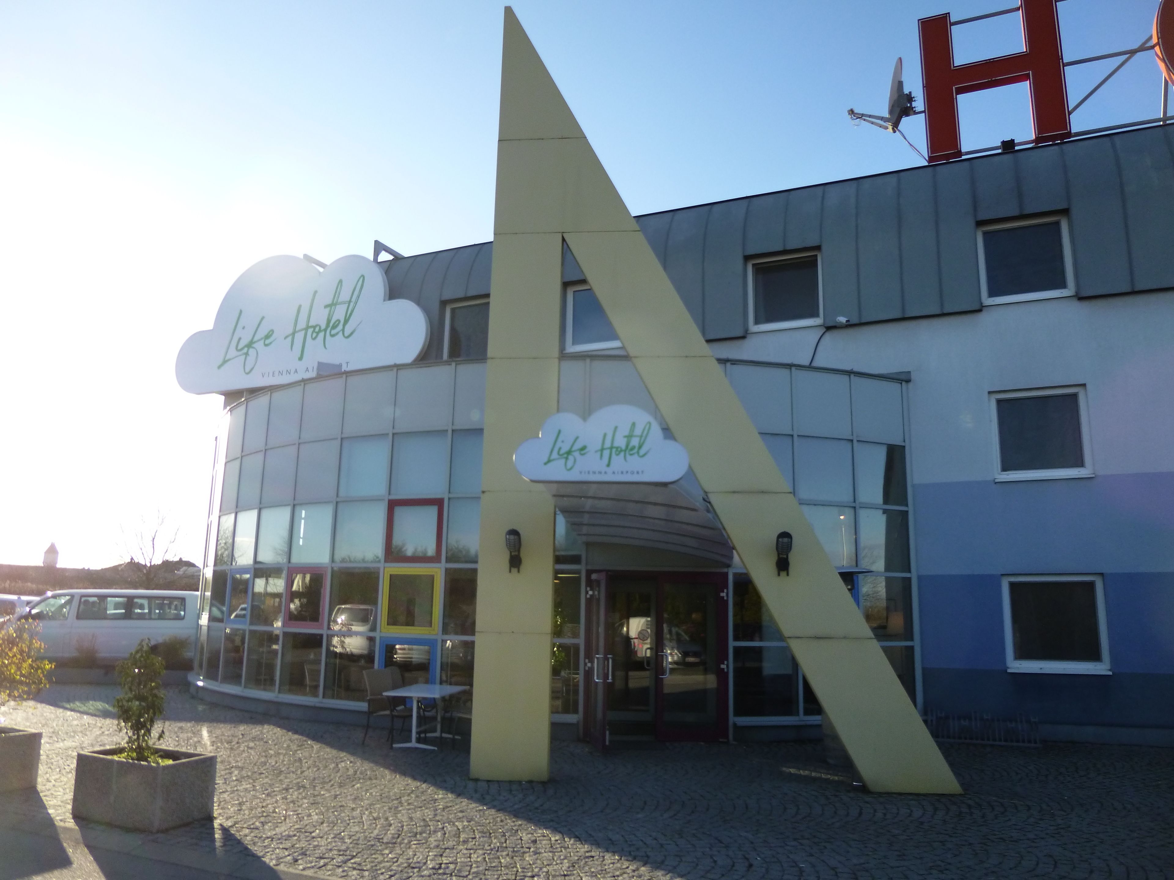 Um hotel com fachada de vidro moderna, um letreiro chamativo 'Life Hotel' e um grande 'H' no telhado.