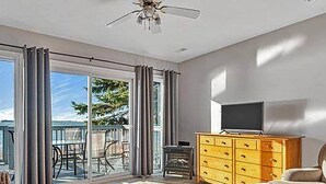 Quarto com sofá, televisão sobre uma cômoda de madeira e varanda com mobiliário externo.